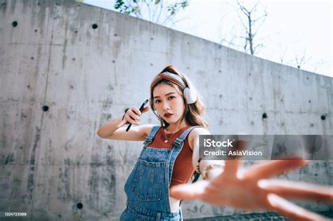 스마트폰을 들고 있는 평온한 젊은 아시아 여성의 초상화가 콘크리트 벽과 햇빛에 맞서 야외에서 헤드폰으로 음악을 들으면서 춤을 추고 있습니다 음악과 라이프스타일 아시아인에