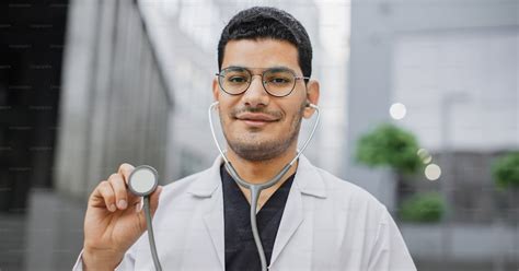 안경을 쓴 친절한 아시아 남성 의사 또는 의대생이 현대 병원 밖에 서서 청진기를 카메라에 시연하는 초상화 얼굴에 초점 맞추기 사진 Unsplash의 아랍 남자 이미지