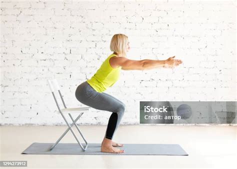 의자가있는 요가 로프트 화이트 스튜디오 실내 선택적 초점에서 매트에 소품과 성인 백인 여성 연습 스쿼트를 맞춥니다 의자에 대한