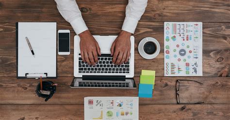 노트북에 입력하는 인간의 손의 상위 뷰입니다 노트북 스마트폰 일기 커피 컵 안경 차트 및 갈색 나무 책상에 있는 기타