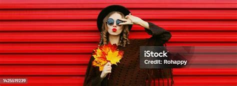 노란색 단풍 나무 잎을 가진 아름다운 젊은 여성의 가을 초상화는 갈색 니트 판초를 입은 립스틱으로 입술을 불고 빨간색 배경에 검은 둥근 모자를 씁니다 패션에 대한 스톡 사진