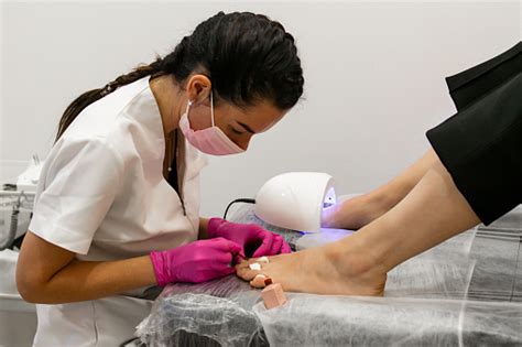 의학 마스크의 페디큐어 마스터는 바니시로 발톱을 페인팅합니다 고전적인 페디큐어를 받는 스파 살롱에서 여자 다리 다리와 손톱 스킨 케어 Spa 절차 2명에 대한 스톡 사진 및