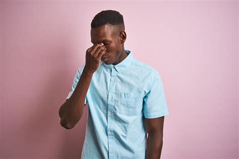 고립 된 분홍색 배경에 서있는 파란색 캐주얼 셔츠를 입은 아프리카 계 미국인 남성이 피로와 두통을 느끼는 코와 눈을 문지르는