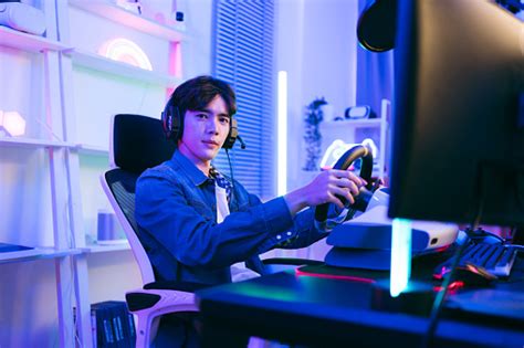 청바지 재킷을 입은 젊은 아시아 남자 프로게이머가 Pc 키보드 모니터 마이크가 있는 게임 테이블이 있는 의자에 앉아 있다 경쟁을 준비하고 게임 플레이를 캐스팅하거나