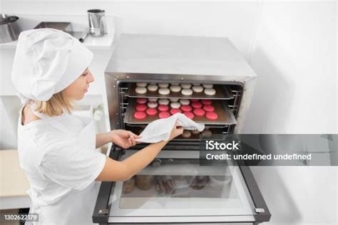 제과 점 마카롱 베이킹 흰색 유니폼과 모자를 쓴 예쁜 여성 과자는 베이킹 트레이에 마카롱 을 배치하여 베이킹 후 오븐에서 꺼낼 준비를합니다 가정 주방에 대한 스톡 사진 및