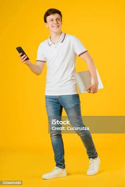 흰색 티셔츠를 입은 젊은 행복한 남자의 초상화가 노란 배경에 노트북 컴퓨터와 스마트폰을 들고 미소짓고 있습니다 20 29세에 대한 스톡 사진 및 기타 이미지 Istock