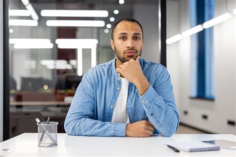 데님 셔츠를 입고 책상에 앉아 손으로 머리를 받치고 카메라를 바라보는 사려 깊은 젊은 무슬림 회사원의 초상화 남자 사업가에 대한 스톡 사진 및 기타 이미지 Istock