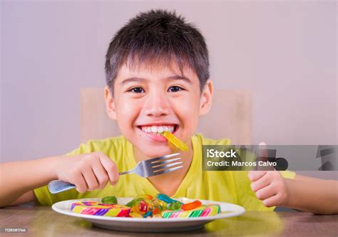 젊은 행복 하 고 흥분 남자 아이 명랑 먹는 접시 가득 사탕과 막대 사탕 설탕 중독 및 남용과 달콤한 건강에 해로운 영양에 회색 배경에 고립 된 테이블에 앉아 웃 고 건강에