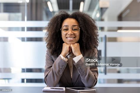 양복을 입고 책상에 앉아 손을 턱 밑에 넣고 카메라를 바라보는 미소 짓는 아프리카계 미국인 여성 사무원의 클로즈업 사진 갈색 머리에 대한 스톡 사진 및 기타 이미지 Istock
