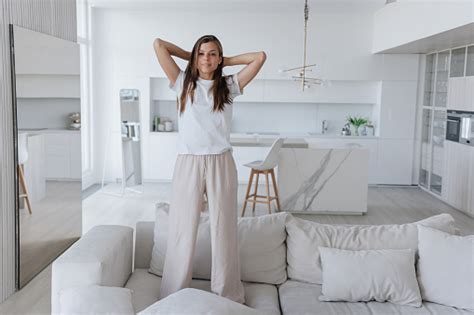 흰색 티셔츠와 베이지색 바지를 입은 독립적인 갈색 머리 젊은 성인 여성이 아늑한 소파에 서서 머리 뒤쪽에 손을 얹고 카메라를 바라보고 있다 호텔에서 휴식을 취하는 목적이