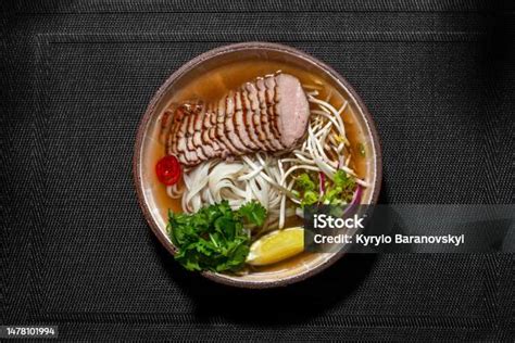 송아지 고기 고수 국수 칠리 잘게 썬 파 마이크로그린 콩나물 라임 웨지를 곁들인 아시아 라면 수프 둥글고 깊고 갈색의 세라믹 그릇에 수프 그릇은 검은 고리 버들 세공 스탠드