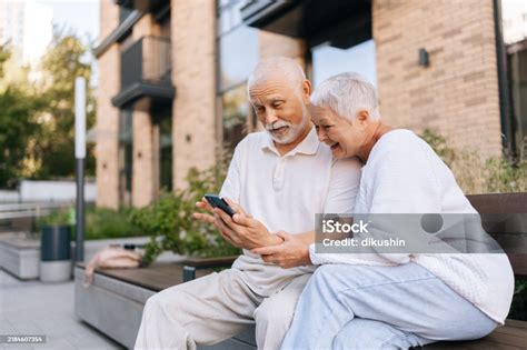 긍정적 인 노인 커플은 도시 벤치에 앉아서 포옹하고 행복하게 스마트 폰을 사용하고 함께 인터넷을 검색하는 시간을 즐깁니다 봄에 대한 스톡 사진 및 기타 이미지 Istock