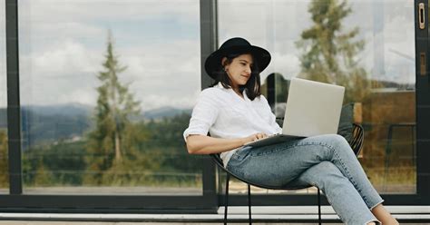 세련된 힙스터 소녀는 큰 창문의 테라스에 노트북을 들고 앉아 있다 노트북을 사용하거나 쇼핑을 하거나 야외에서 집에서 온라인으로 일하는 모자를 쓴 젊은 행복한 여성