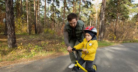 젊은 아버지는 안전 헬멧과 캐주얼웨어를 입은 어린 아들이 공원의 아스팔트 도로를 따라 균형 잡힌 자전거를 타는 것을 도우면서