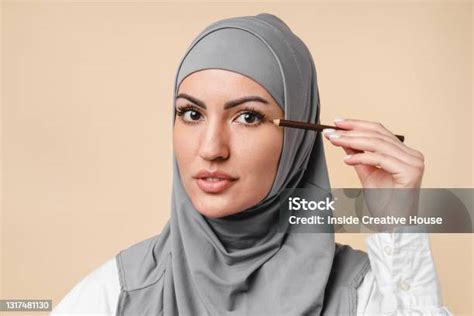 회색 히잡을 입고 있는 예쁜 젊은 아라비아 무슬림 이슬람 여성이 베이지색 배경위에 아이 연필로 메이크업을 하고 있습니다 화장품 깨끗한 피부 바디 케어 미용사 컨셉 뷰티