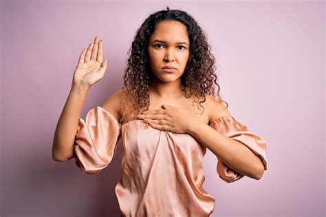 가슴과 열린 손바닥에 손으로 맹세 분홍색 배경 위에 서 캐주얼 티셔츠를 입고 곱슬 머리를 가진 젊은 아름다운 여자 충성 약속 맹세를 만들기 T 셔츠에 대한 스톡 사진 및