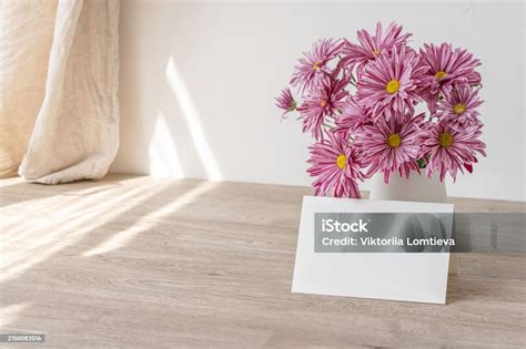 빈 종이 카드 모형 베이지 색 나무 테이블에 분홍색 꽃다발이 달린 꽃병 자연 햇빛 그림자가있는 흰색 벽과 린넨 커튼 배경 미적 인사말 엽서 또는 초대장 템플릿 생일 카드에