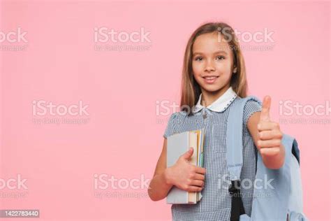 가방과 교과서가 달린 유니폼을 입은 젊은 백인 십대 여학생의 클로즈업 사진 분홍색으로 고립 된 엄지 손가락을 보여주는 카메라를 바라 봅니다 10 11세에 대한 스톡 사진 및