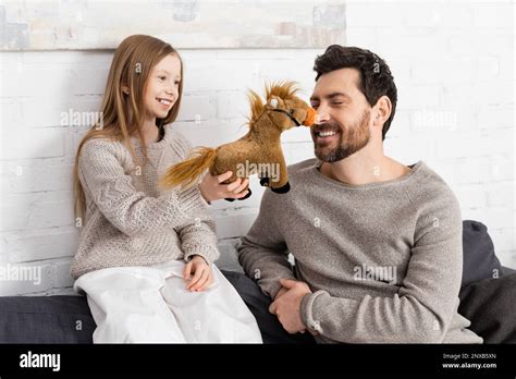 Cheerful Girl Showing Toy Horse To Smiling Dad Sitting On Couch With