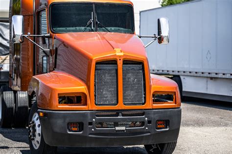 갓 그린 오렌지색 큰 장비 세미 트럭은 프론트 엔드가 분해되어 헤드 라이트와 그릴이 누락 된 상태로 다른 세미 트레일러와 함께 적재 된 세미 트레일러와 함께 일렬로 앉아