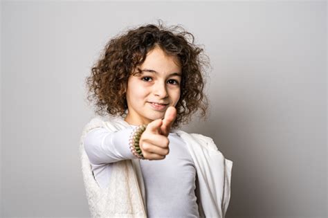 곱슬 머리 앞보기 초상화와 함께 10 살 된 작은 백인 소녀 10 살 은 행복하고 기쁨미소 웃는 손가락 복사 공간을 가리키는 카메라를 찾고 흰색 배경 앞에 서 서 가리키기에