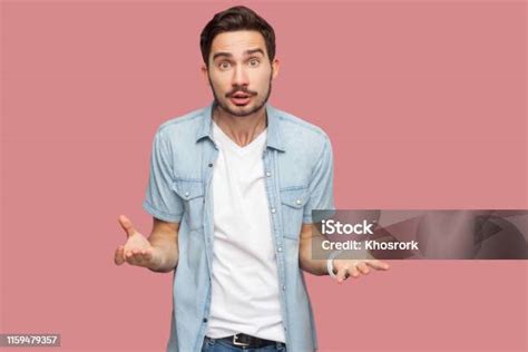 무엇을 원하세요 화가 난 초상화는 파란 캐주얼 스타일의 셔츠를 입고 카메라를 보고 묻는 잘생긴 수염을 가진 젊은이에게 충격을 주었습니다 남자에 대한 스톡 사진 및 기타