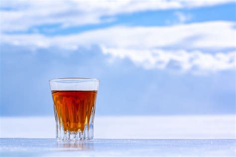 구름이 있는 하늘을 배경으로 얼음판에 술 한 잔이 서 있다 차가운 위스키 유리에 오렌지 음료 공간을 복사합니다 측면 보기입니다 배경이 흐릿합니다 수평 0명에 대한 스톡 사진