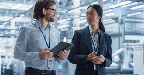 두 명의 다양한 엔지니어가 사무실에서 태블릿 컴퓨터를 사용하여 현대 전자 공장에서 걷고 있습니다 여성과 남성 전문가들이 현대 조립 라인에서 로봇을 모니터링하며 이야기합니다