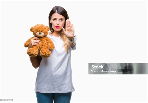 젊은 아름 다운 여 자가 심각 하 고 자신감 표현 방어 제스처 정지 표시를 하 고 오픈 손으로 고립 된 배경 위에 테 디 베어 봉 제를 들고 갈색 머리에 대한 스톡 사진 및