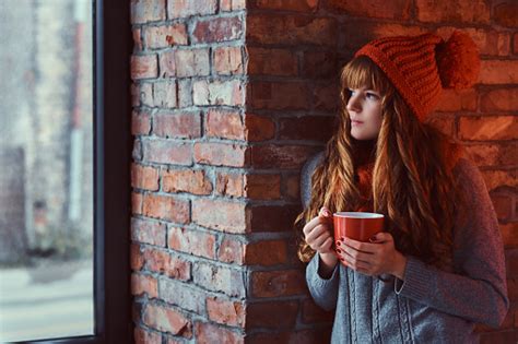따뜻한 스웨터와 벽돌 벽에 기대어 창문을 보고 커피 한 잔을 들고 모자를 입고 빨간 머리 소녀 12월에 대한 스톡 사진 및 기타 이미지 Istock