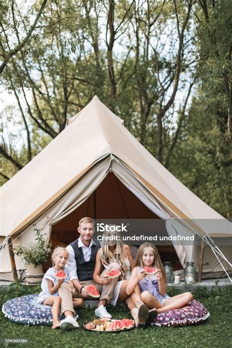 화창한 날에는 공원이나 숲에서 베개에 함께 앉아있는 동안 수박을 먹는 세련된 보헤미안 옷을 입은 행복한 가족 배경에 큰 Wigwam Tipi 텐트 가족에 대한 스톡 사진 및