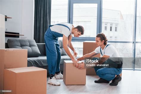상자를 포장합니다 방에서 실내에서 일하는 파란색 유니폼을 입은 두 명의 젊은 이주 이삿짐 옮기는 사람에 대한 스톡 사진 및 기타 이미지 이삿짐 옮기는 사람 새 집으로
