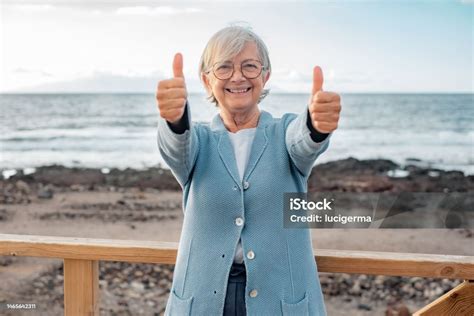 푸른 옷을 입은 웃는 노인 여성이 일몰 해변에서 야외에 서서 엄지손가락을 치켜들고 카메라를 보고 있다 기쁨과 긍정을 표현하는 행복한 연금 수급자 여자 경관에 대한 스톡 사진