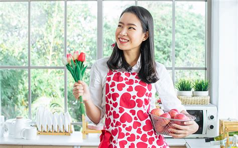 아름다운 아시아 여성은 심장 패턴으로 앞치마를 입고 행복으로 웃고 꽃을 들고 빨간 사과 바구니를 들고 발렌타인 데이에 집에서 부엌에 서 있습니다 라이프 스타일 사랑 과일