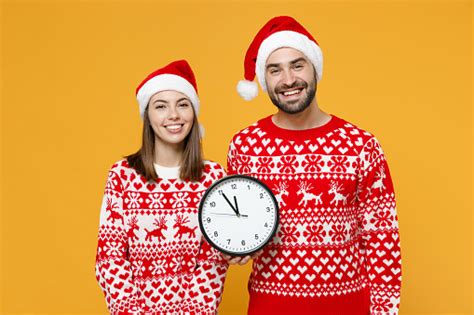 미소 짓는 쾌활한 젊은 산타 커플 친구 남자 여자 20 S 입고 빨간 스웨터 크리스마스 모자 보유 시계 고립 된 노란색 배경 스튜디오 초상화 새해 축하 메리 홀리데이 컨셉