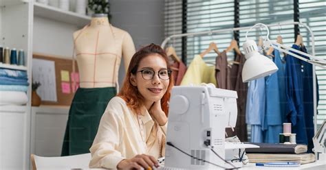 아시아의 젊은 재단사 소녀는 아틀리에를 재단하고 카메라를 봅니다 매력적인 젊은 여성 패션 디자이너는 작업장에서 새로운 수제 양복을 만드는 재봉틀을 통해 직물 재료를