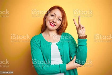 Youg 아름다운 빨간 머리 여자 겨울 녹색 스웨터 위에 고립 된 노란색 배경 에 행복한 얼굴 윙크 와 함께 미소 승리 기호를하고 2번 긍정적인 감정 표현에 대한 스톡 사진