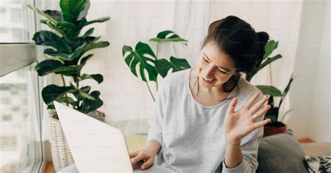 노트북에 인사를 하고 가족이나 친구와 영상 채팅을 하고 베개와 식물이 있는 현대적인 방에 앉아 있는 행복한 소녀 격리 구역에서 통신을 위해 노트북을 사용하는 젊은