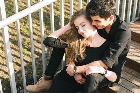 아름 다운 젊은 여러 인종 커플 사랑에 학생 부부는 도시에서 나무 계단을 앉아 있다 아름 다운 터키어 갈색 머리 남자가 검은 옷에 백인 여자를 포옹 화창한 날씨에 젊은이