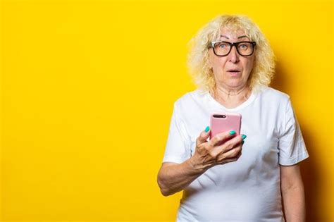 Удивленная блондинка старуха в белой футболке и очках держит телефон