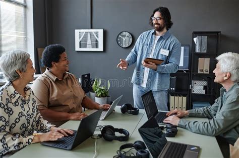 Bearded Young Man Leading Computer Class For Seniors Using Laptops At