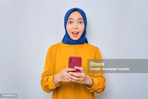 오렌지색 셔츠를 입고 휴대폰을 들고 흰색 배경에 고립된 카메라를 보고 충격을 받은 젊은 아시아 무슬림 여성 20 24세에 대한 스톡 사진 및 기타 이미지 Istock