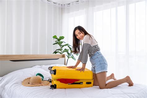 행복한 젊은 아시아 여성은 짐을 포장하고 미소를 지으며 짐을 닫으려고노력합니다 여름 방학 여행 여행을위한 가방을 준비하는 소녀 카메라를보고 20 29세에 대한 스톡 사진 및