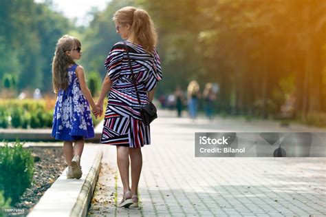 햇볕이 잘 드는 여름 공원 골목에서 손을 잡고 걷는 선글라스와 세련된 드레스를 입은 젊은 금발의 긴 머리 의 슬림 한 매력적인 여성과 어린 소녀의 뒷모습 행복한 가족 관계
