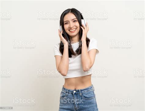 쾌활한 아시아 여성 십대는 고립된 흰색 배경에 흰색 헤드폰 춤을 추며 음악을 듣습니다 손에 아름 다운 젊은 여자는 음악과 함께