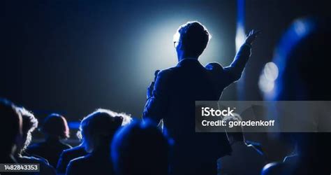 스타트업 서밋의 어둡고 붐비는 강당에 있는 스타일리시한 젊은 사업가의 뒷모습 Qa 세션 중에 마이크에 대고 이야기하는 젊은이 이벤트 연사와 함께 행복한 기업가 컨퍼런스