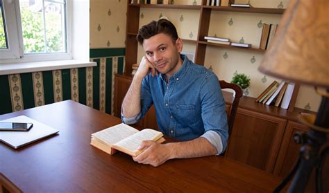 데님 셔츠를 입은 젊은 남자가 나무 책상에 앉아 손에 머리를 기대고 책을 읽고 있습니다 스마트폰과 노트북이 근처에 있으며 배경에는 자연 채광과 책장이 있습니다 30 34세에