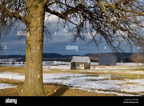 One Room Log Cabin Shack With Solar Panel In Farmers Field With Snow Clouds And Naked Tree In