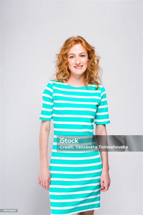 회색 배경에 스튜디오에서 파란색 줄무늬가 있는 여름 드레스에 빨간 곱슬 머리를 가진 젊고 아름 다운 여자의 무릎 초상화 여름 방학 관광 및 여름 옷의 테마 관광객에 대한