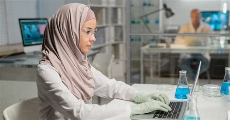 히잡 실험실 코트 보호 안경 장갑을 낀 젊은 무슬림 여성이 실험실에서 노트북 앞에 책상 옆에 앉아 타이핑을 하고 있다 사진